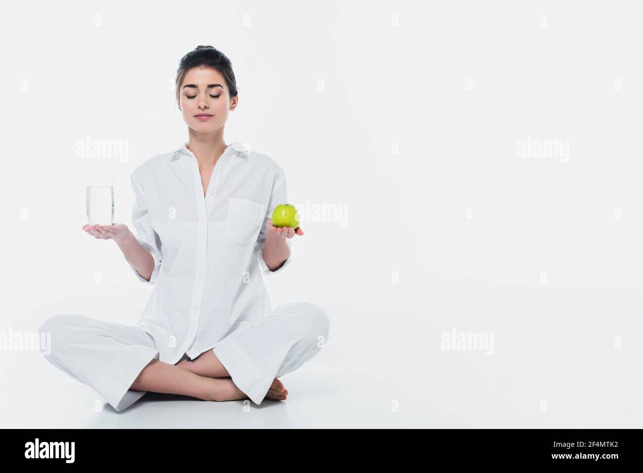 Brunette woman with glass of water and apple sitting in yoga pose on ...