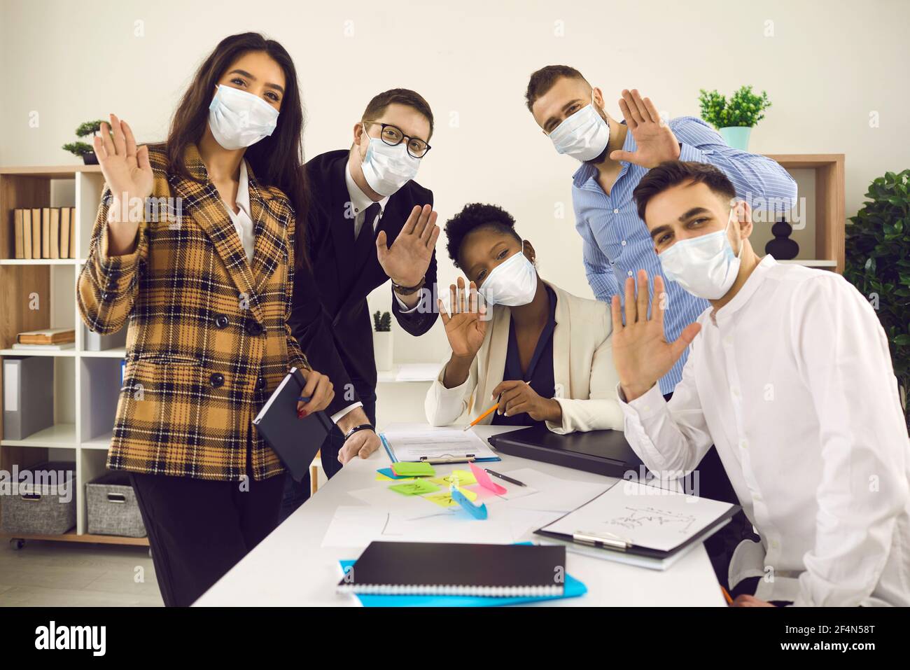 Happy creative office worker team in face mask waving hello to camera ...