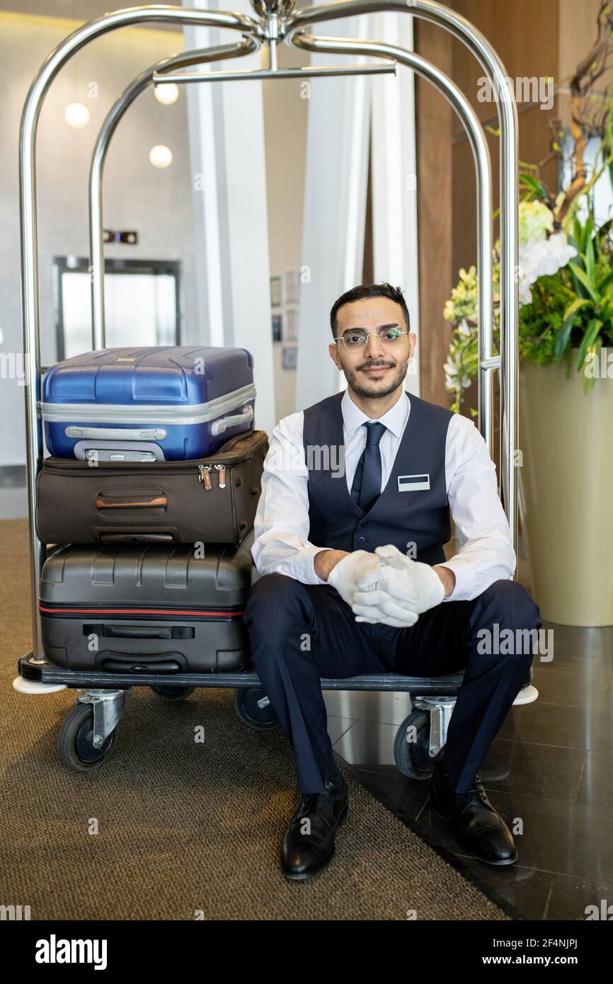 Happy young porter in uniform, gloves and eyeglasses sitting on cart ...