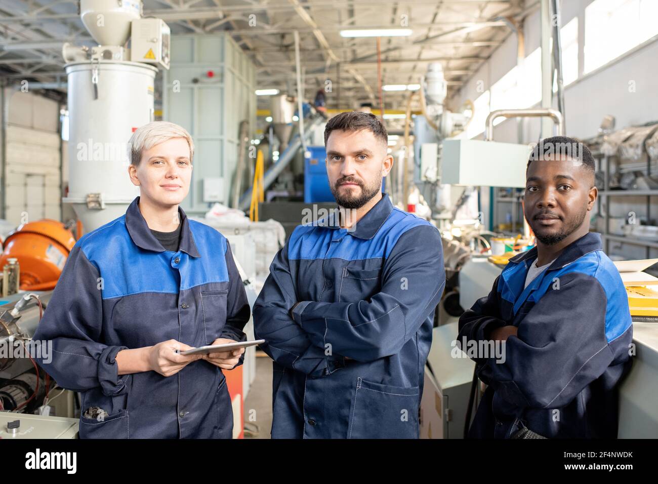 Three young serious workers of contemporary plant in overalls standing ...