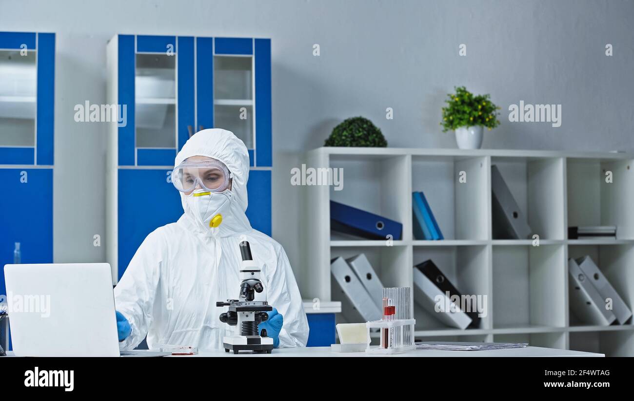 scientist in hazmat suit working near laptop and microscope in ...