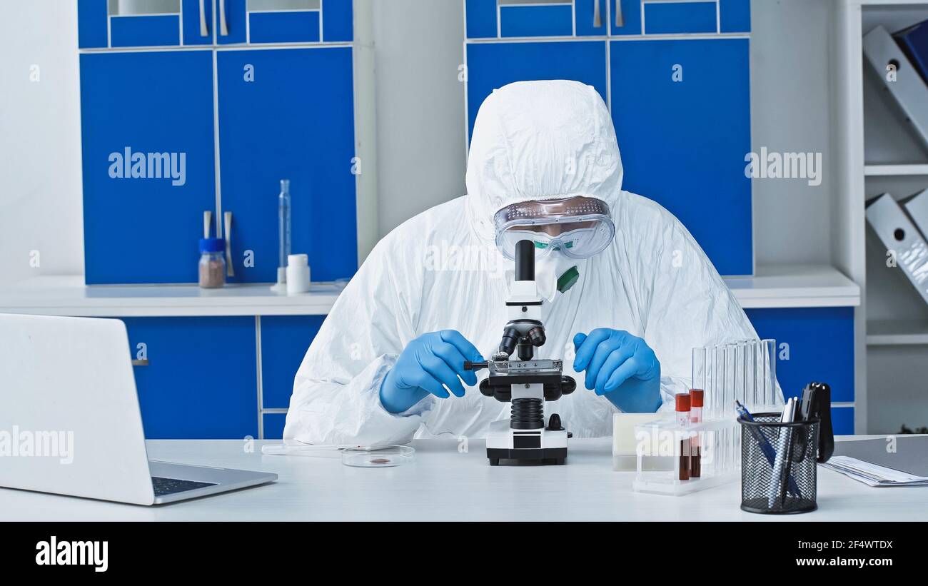 virologist in hazmat suit doing working with microscope near laptop in ...