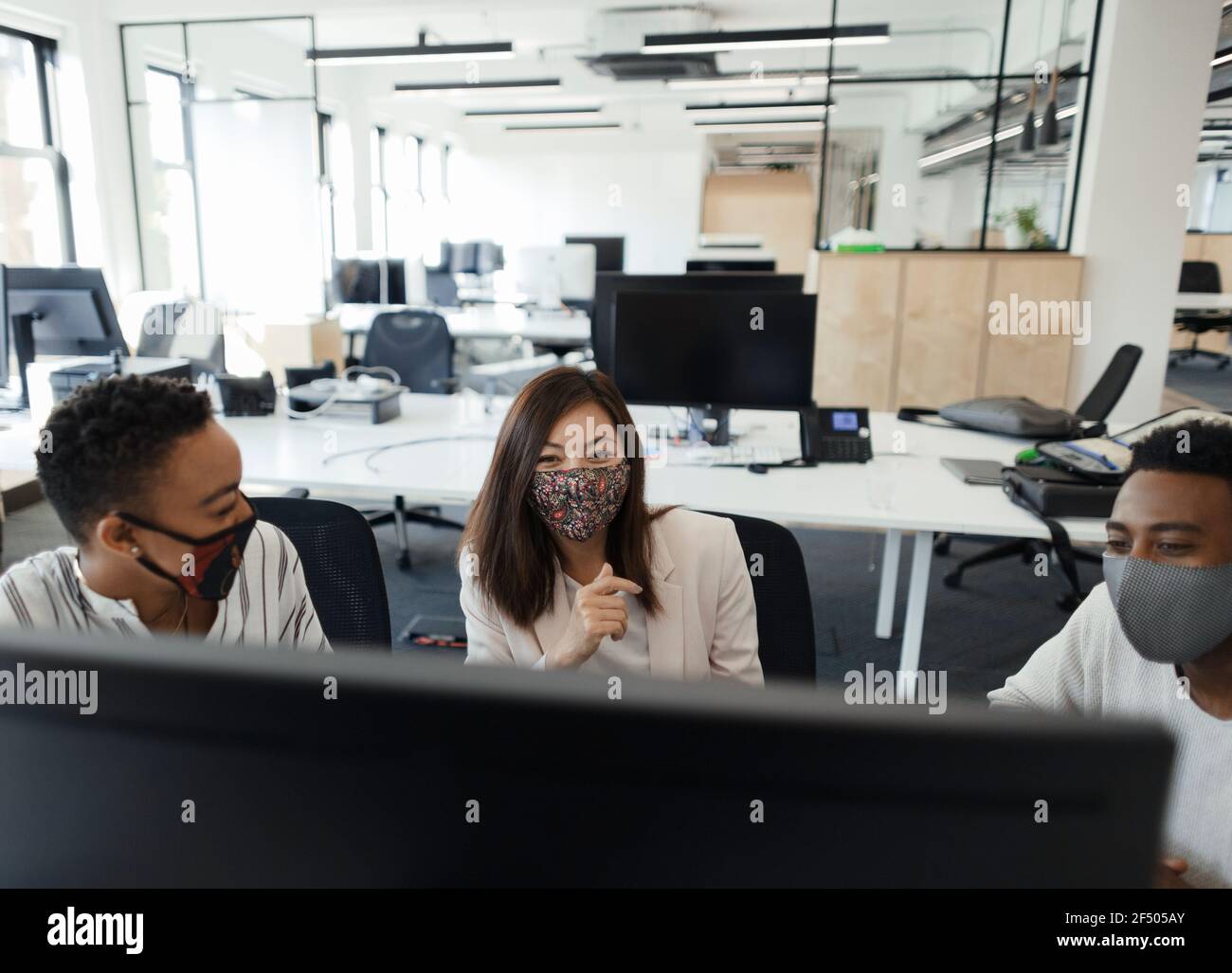 Business people in face masks working at computer in open plan office ...