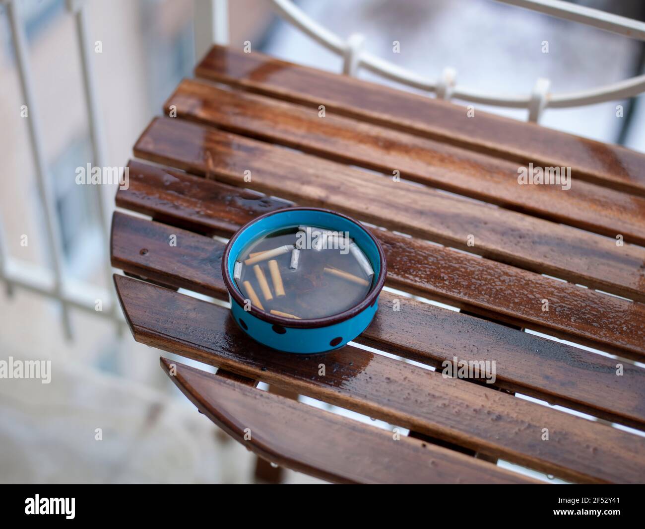 Ashtray full of water placed on a wet wooden table top, outdoor closeup ...