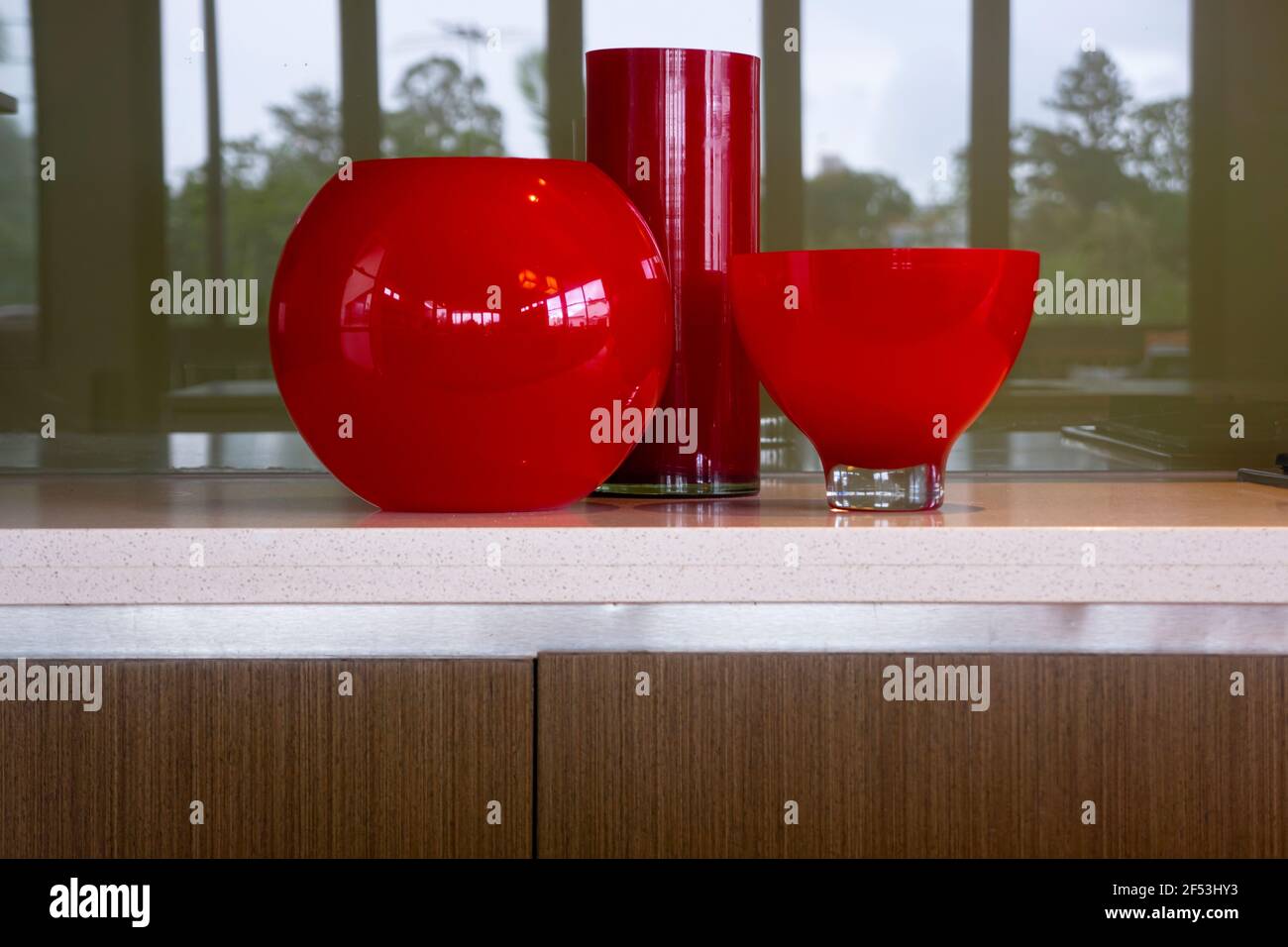 Three bright red vases on a kitchen bench with a green glass background ...
