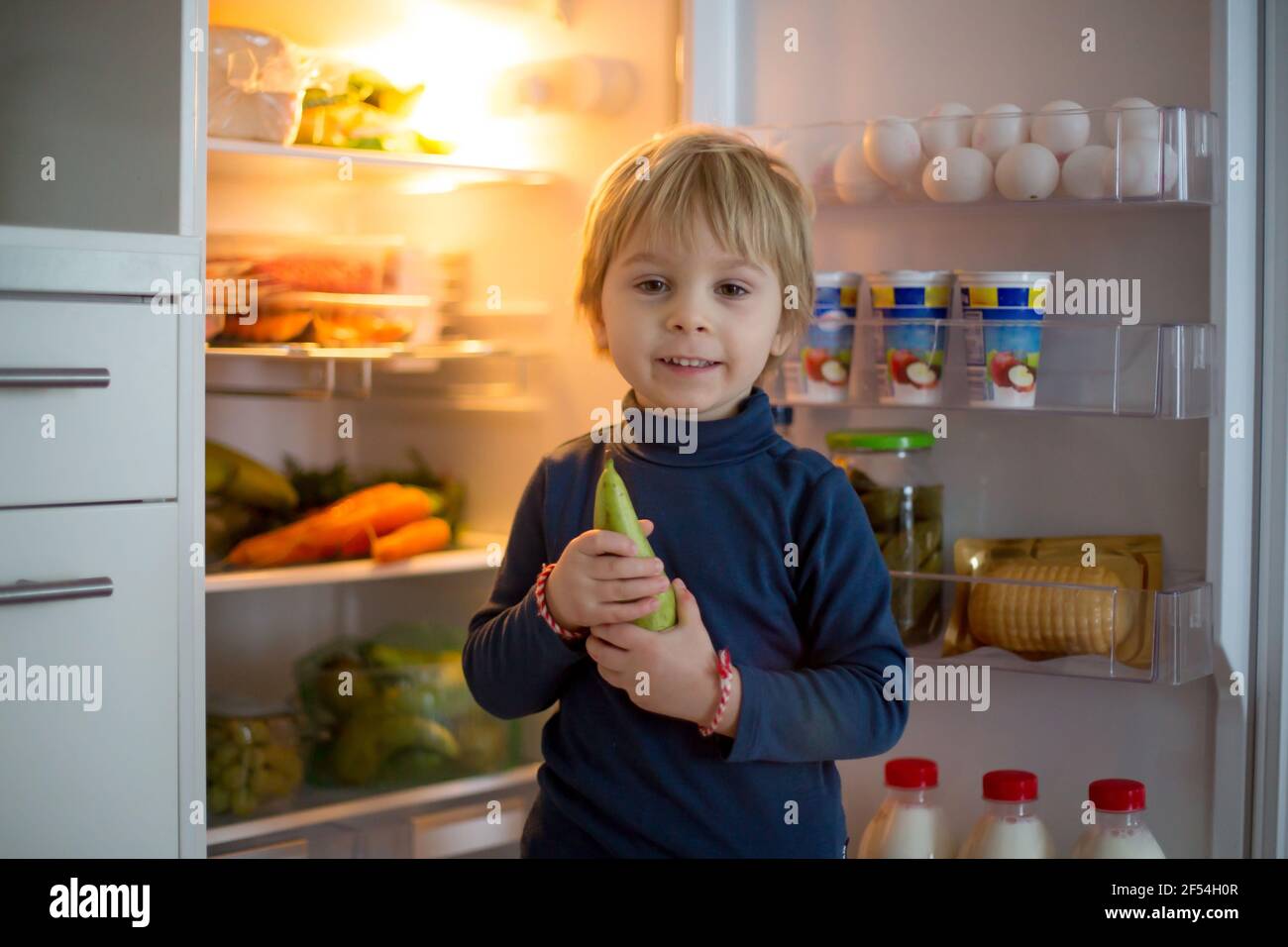 Cute toddler blond child, opening the fridge door and taking fruits adn ...
