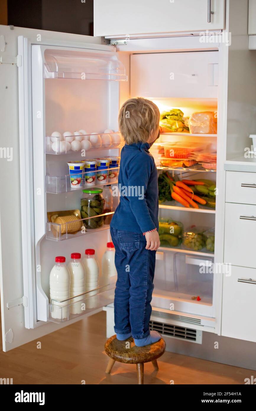 Cute toddler blond child, opening the fridge door and taking fruits adn ...