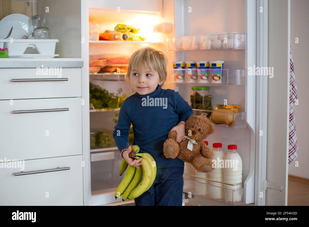 Cute toddler blond child, opening the fridge door and taking fruits adn ...