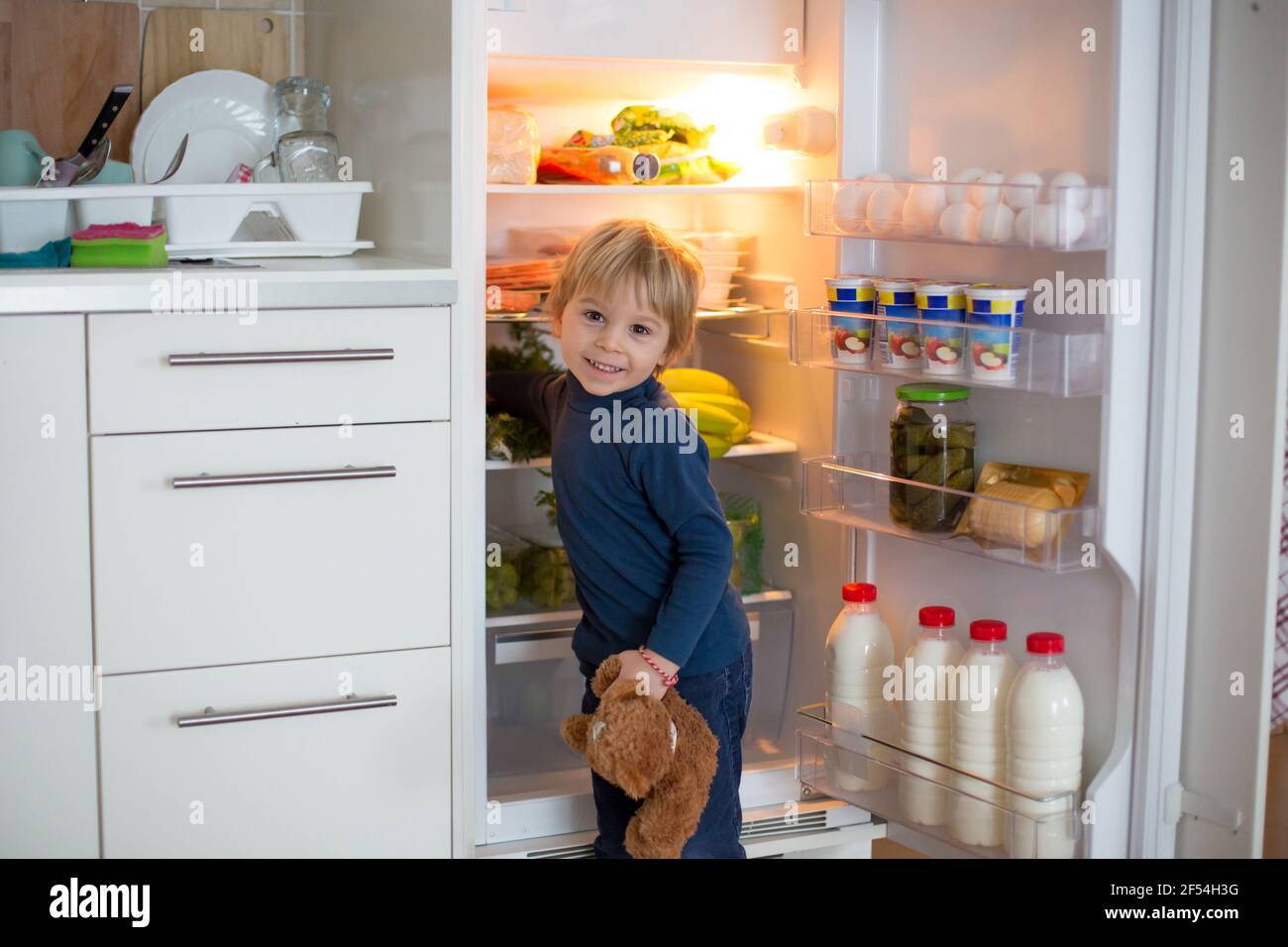 Cute toddler blond child, opening the fridge door and taking fruits adn ...
