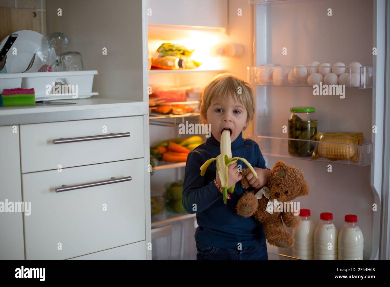 Cute toddler blond child, opening the fridge door and taking fruits adn ...