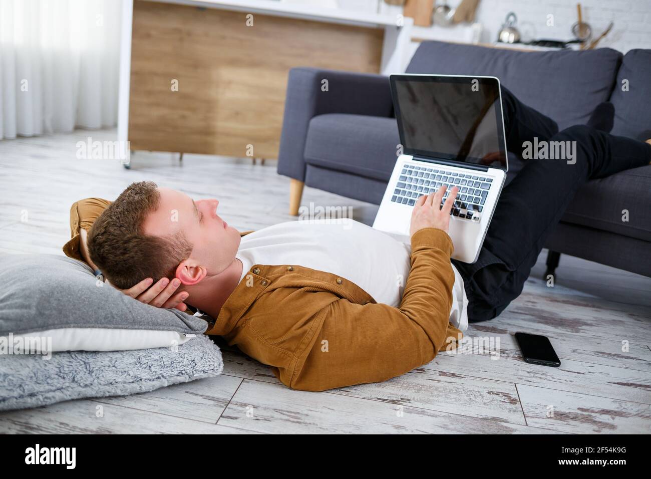 Attractive smiling young man in casual wear sitting on floor on sofa in ...