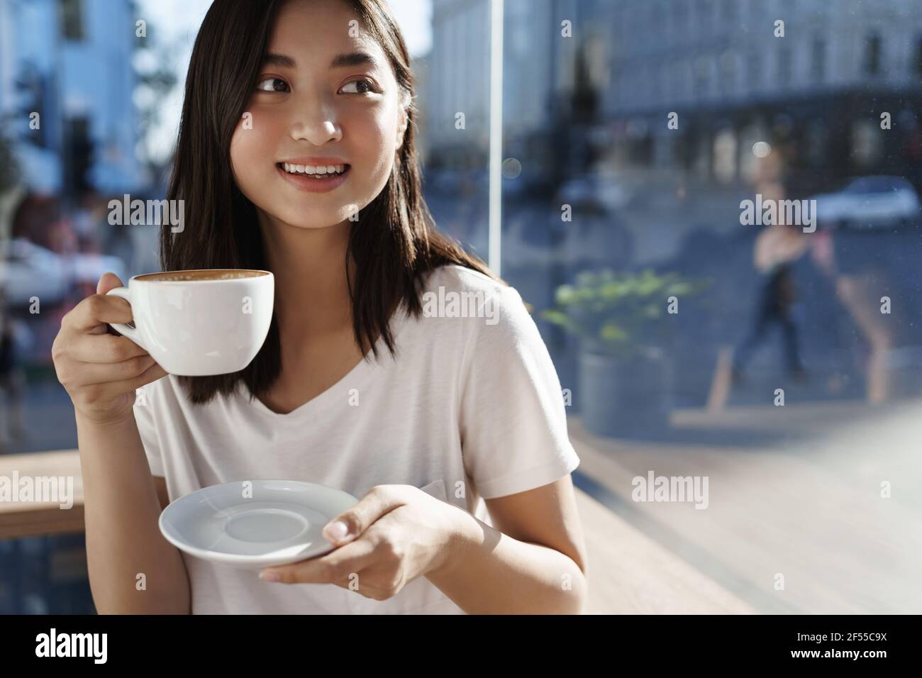 Modern woman sitting in cafe near window and looking outside, drinking ...