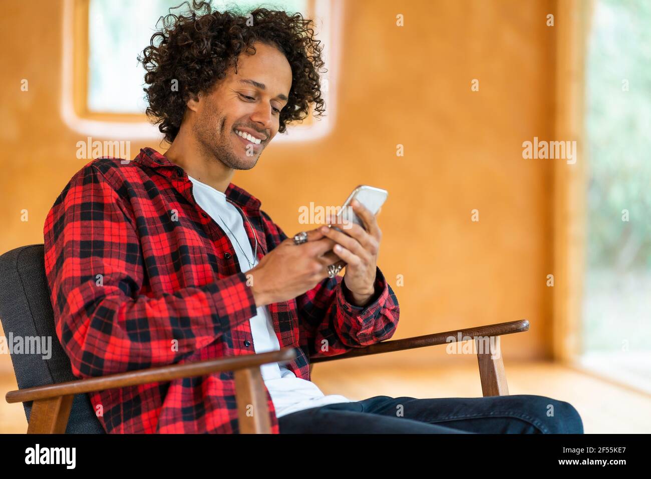 Young man using smart phone while sitting on armchair at spacious room ...