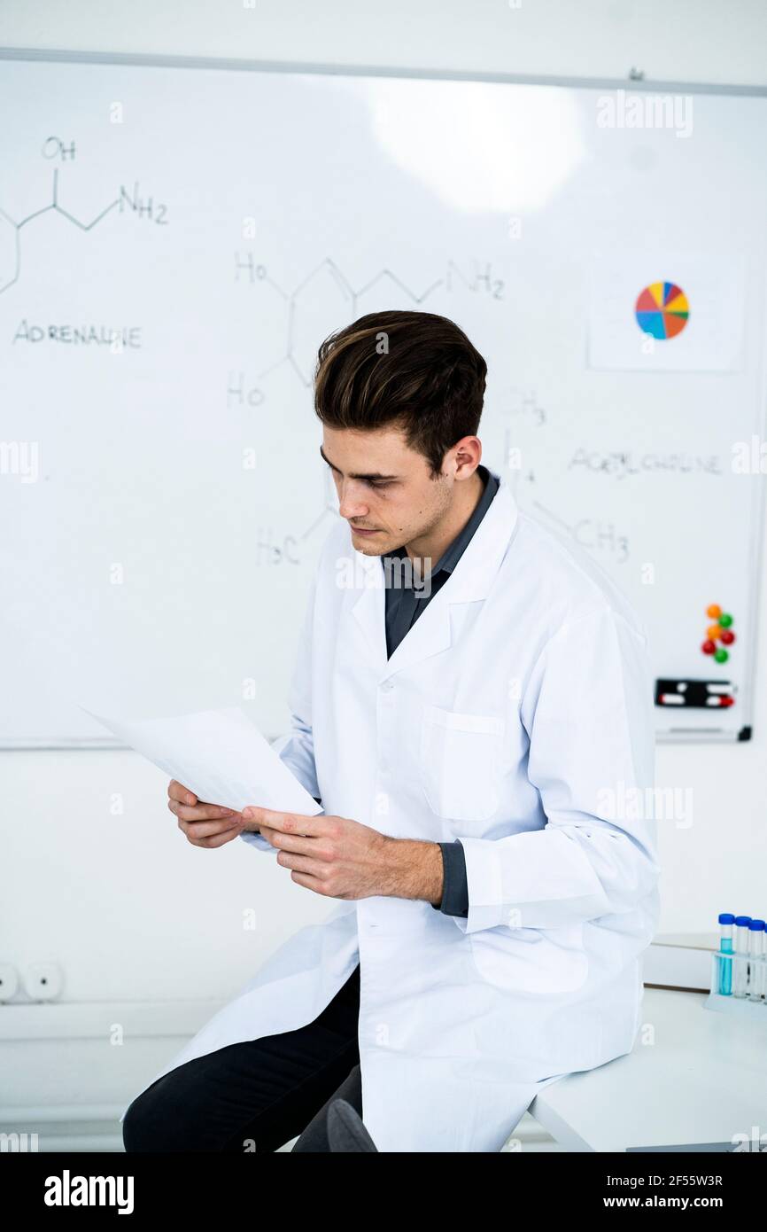 Young male scientist looking at chart while sitting on desk in ...