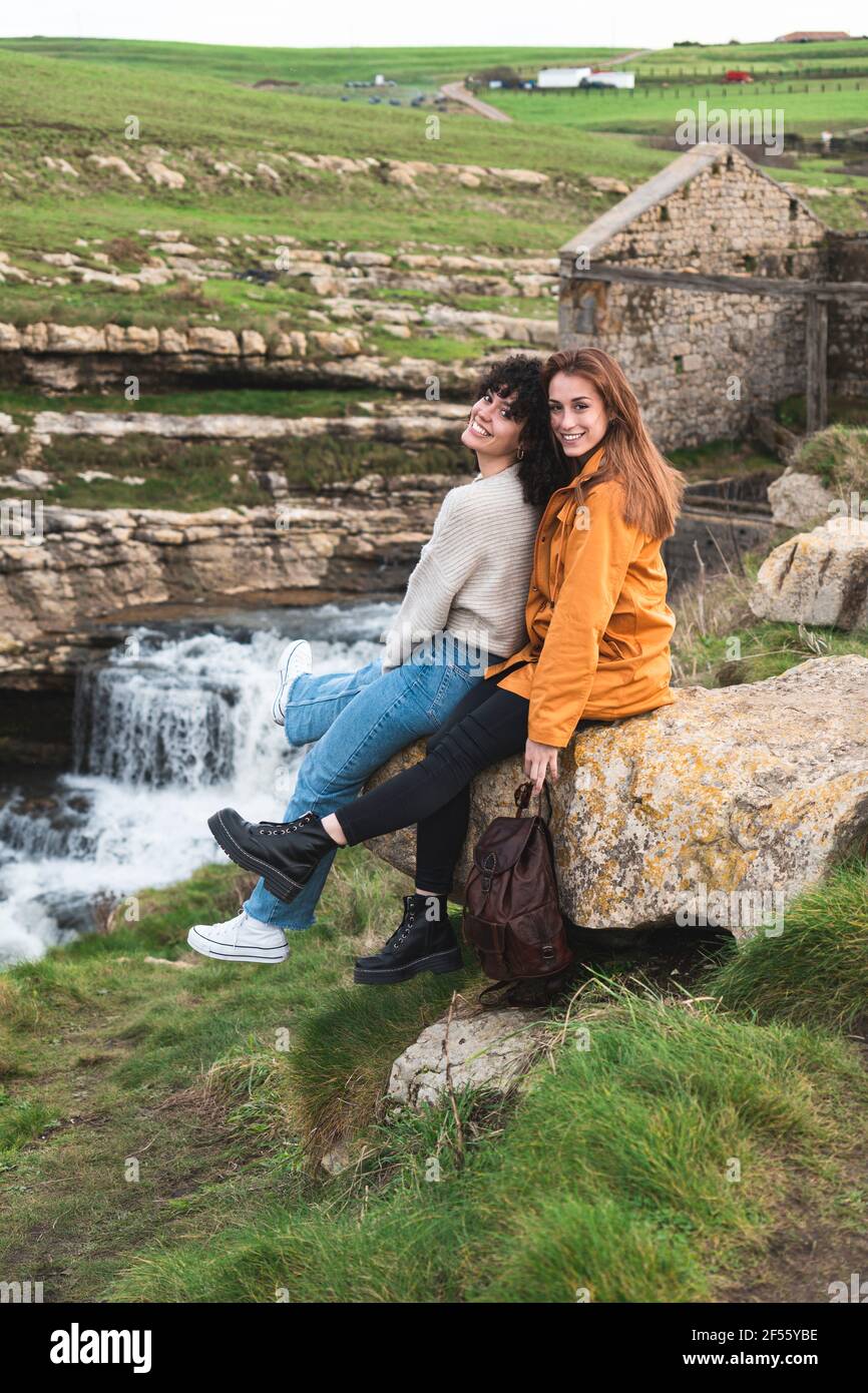Happy female friends sitting on rock by waterfall Stock Photo - Alamy