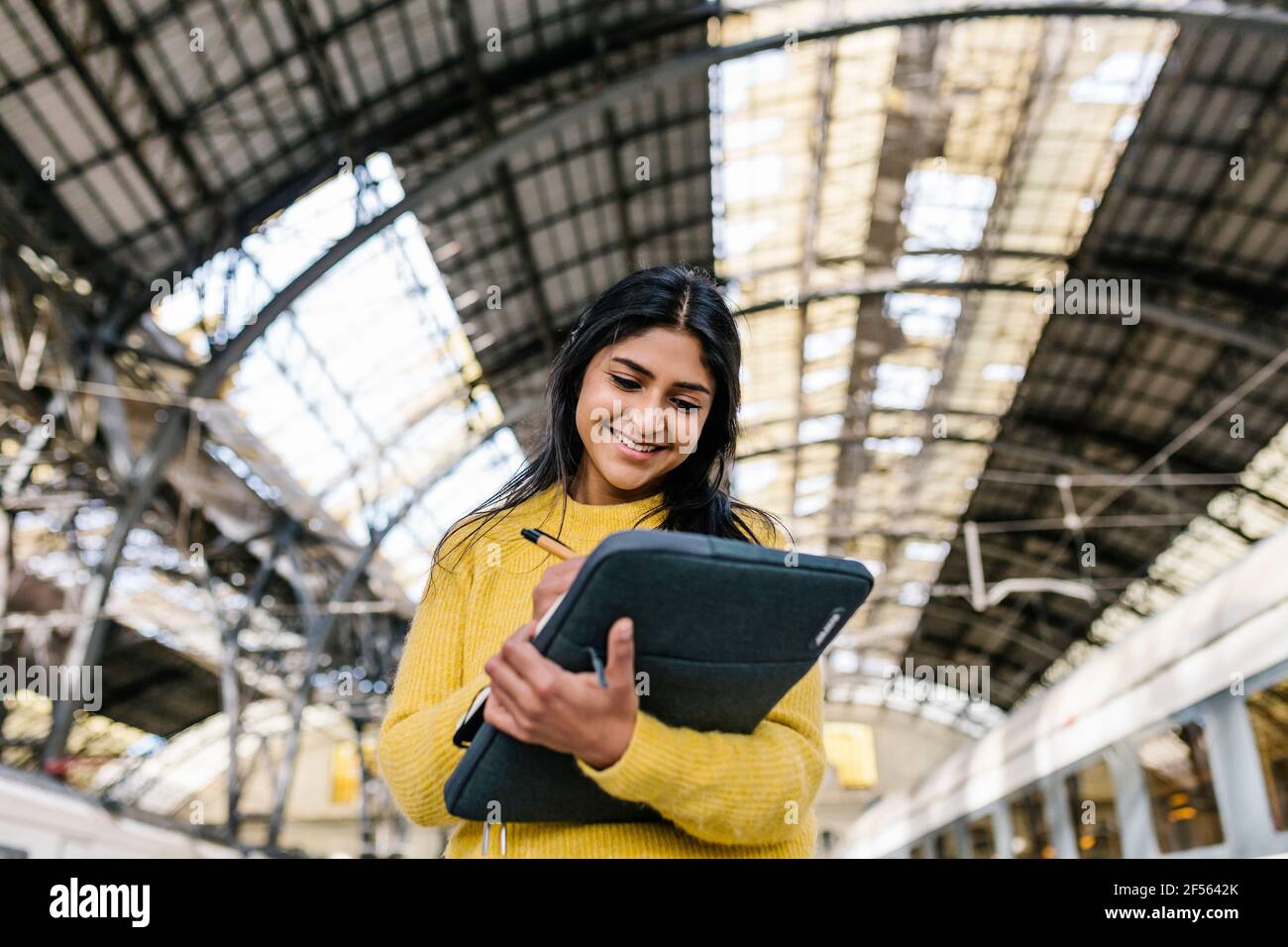 Smiling woman with laptop bag writing in diary on railroad station ...