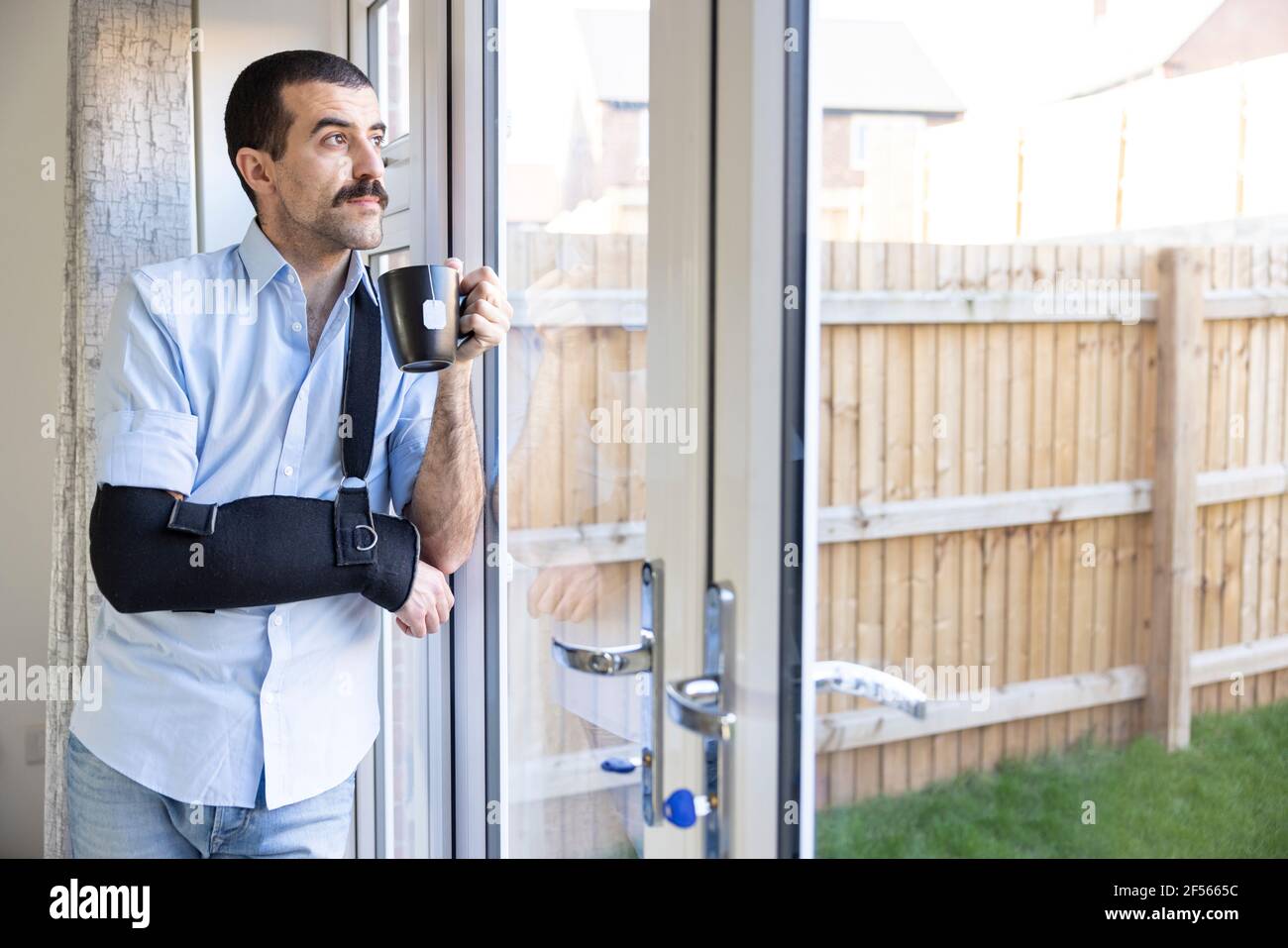 Thoughtful man wearing arm sling drinking tea while looking through ...