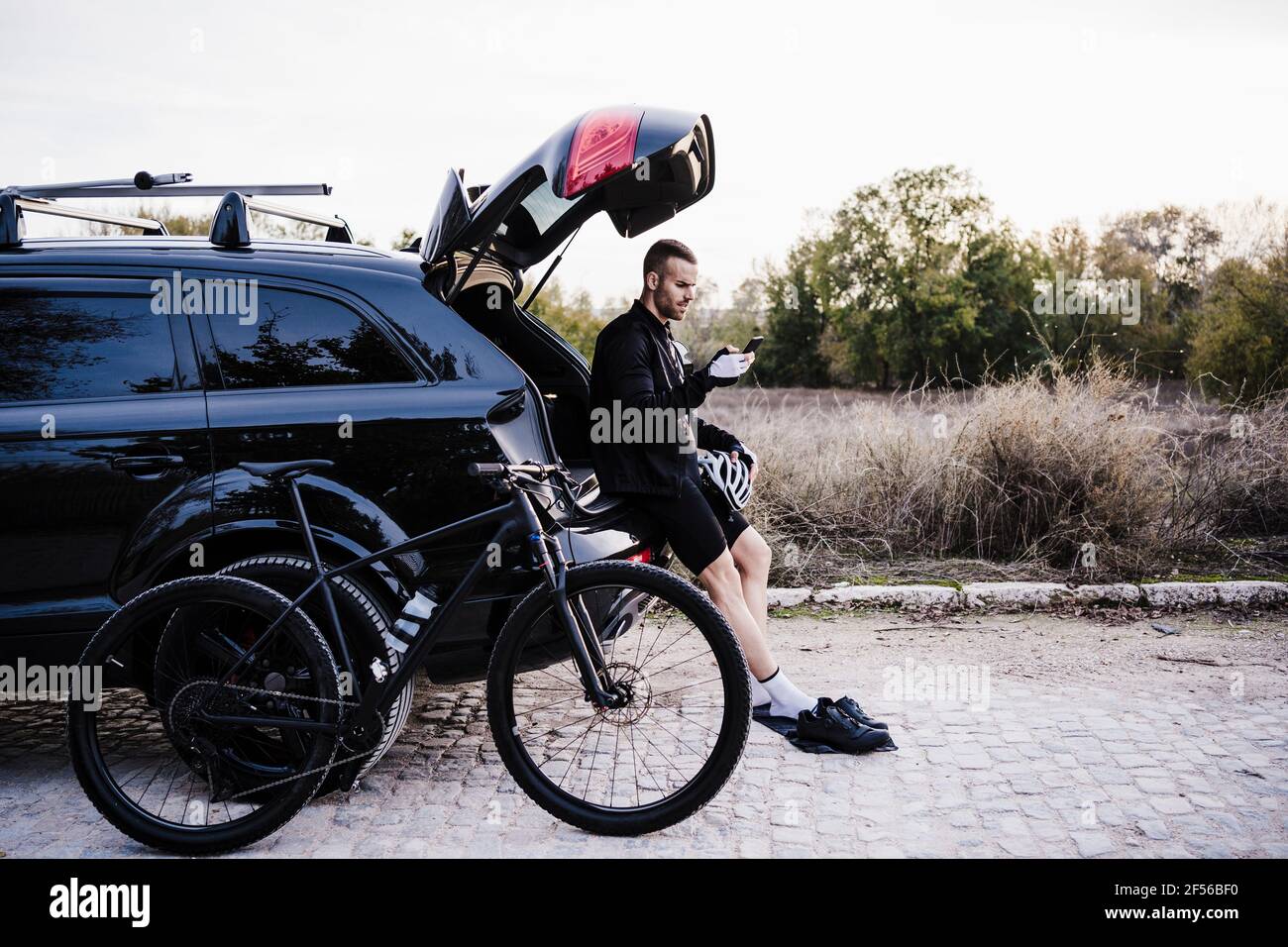 Male cyclist using mobile phone while sitting in car trunk by bicycle ...