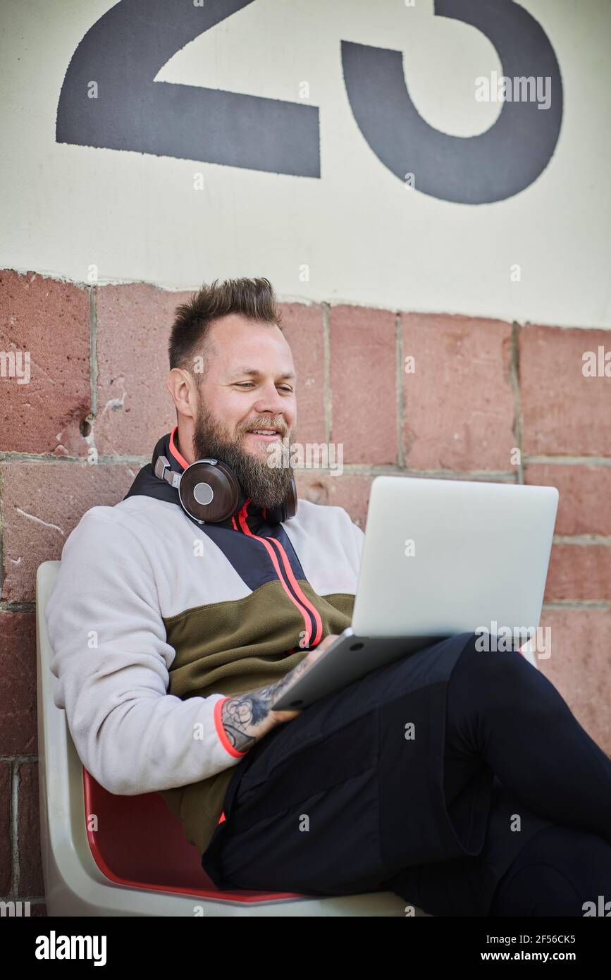 Smiling male coach using laptop while sitting on bench against wall ...
