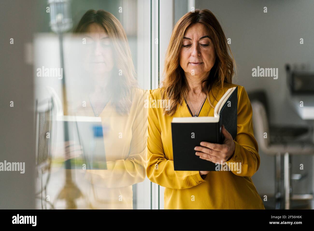 Woman reading book while leaning on glass window at home Stock Photo ...