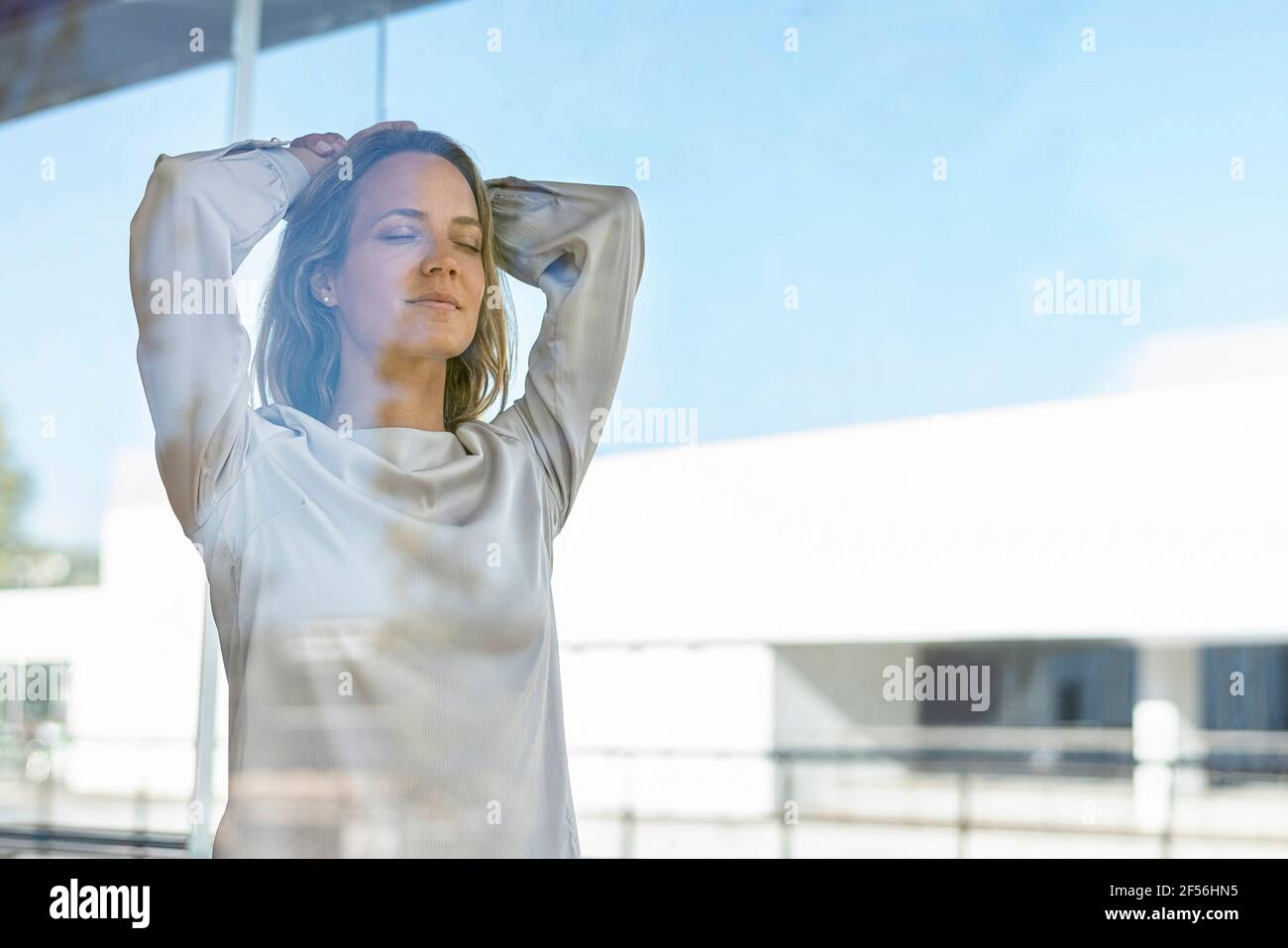 Businesswoman with hands behind head and eyes closed standing by glass ...