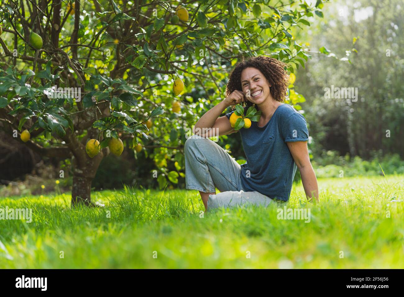 Happy woman holding lemons while sitting on grass under lemon tree ...