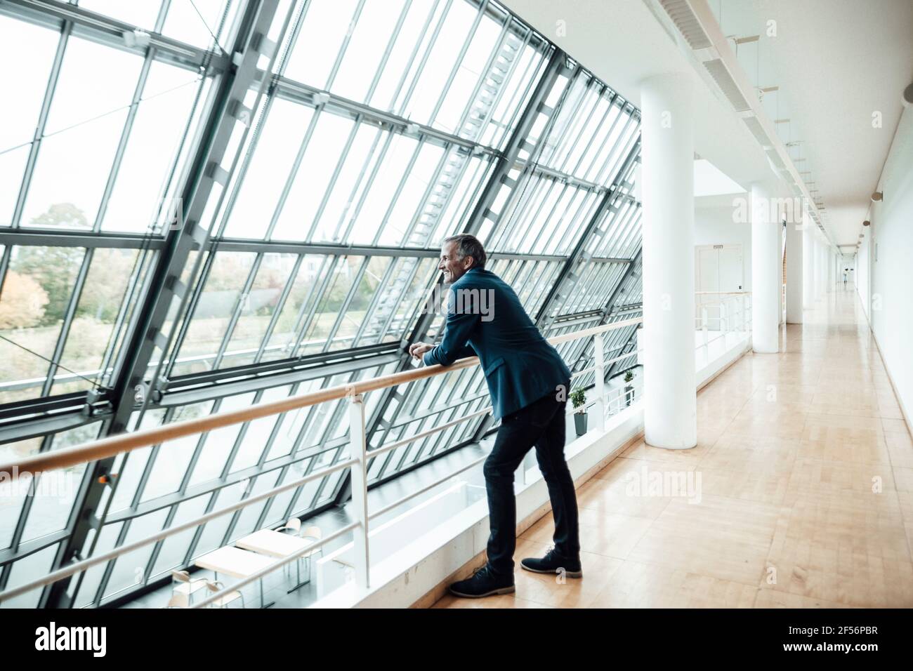 Smiling businessman looking though glass while leaning on railing in ...