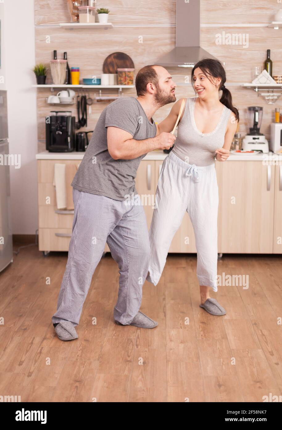 Joyful couple dancing and singing during breakfast in kitchen wearing ...
