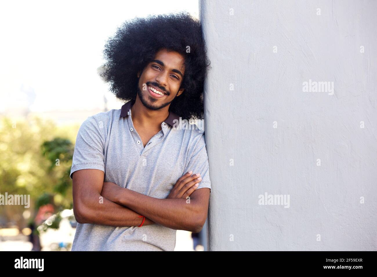 Portrait of young man standing with arms crossed leaning on white wall ...