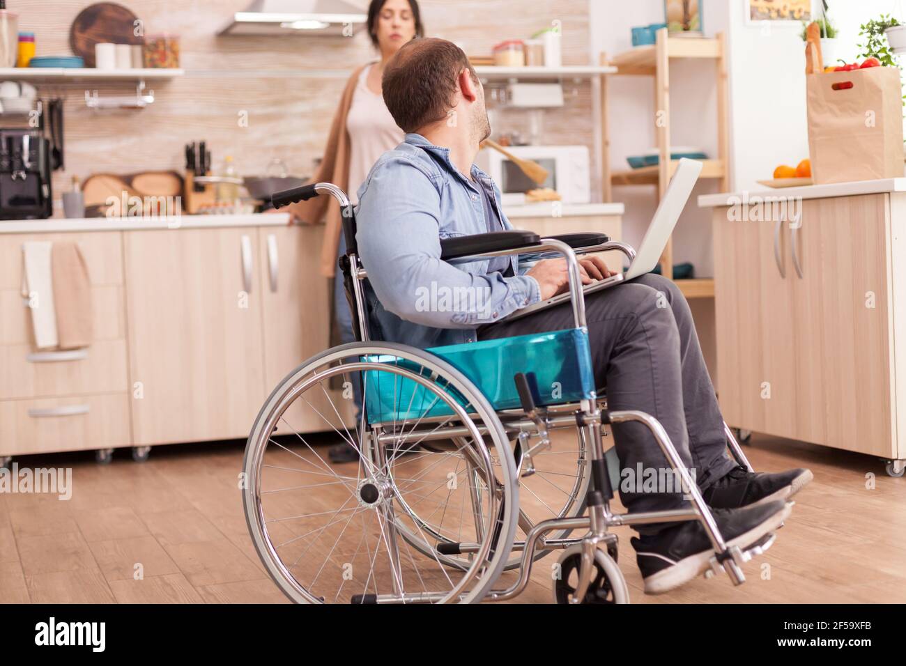 Disabled man in wheelchair working on laptop while wife is cooking in ...