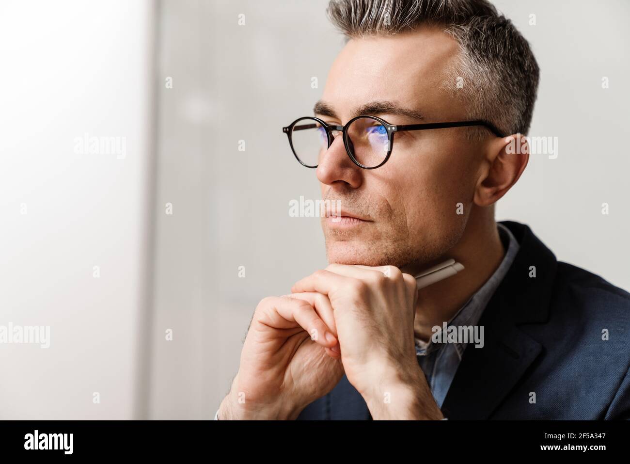 Thinking grey man wearing eyeglasses holding pen and looking forward in ...