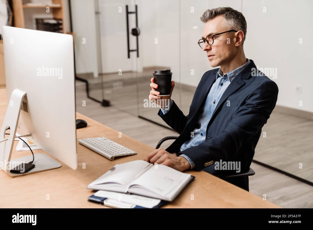 Focused grey man drinking coffee while working with computer in office ...