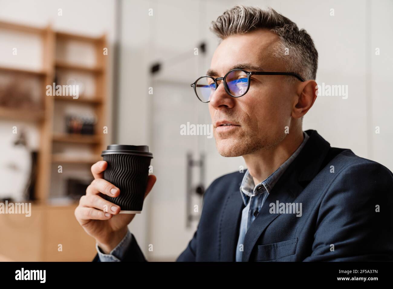 Focused grey man in eyeglasses drinking coffee while working at office ...