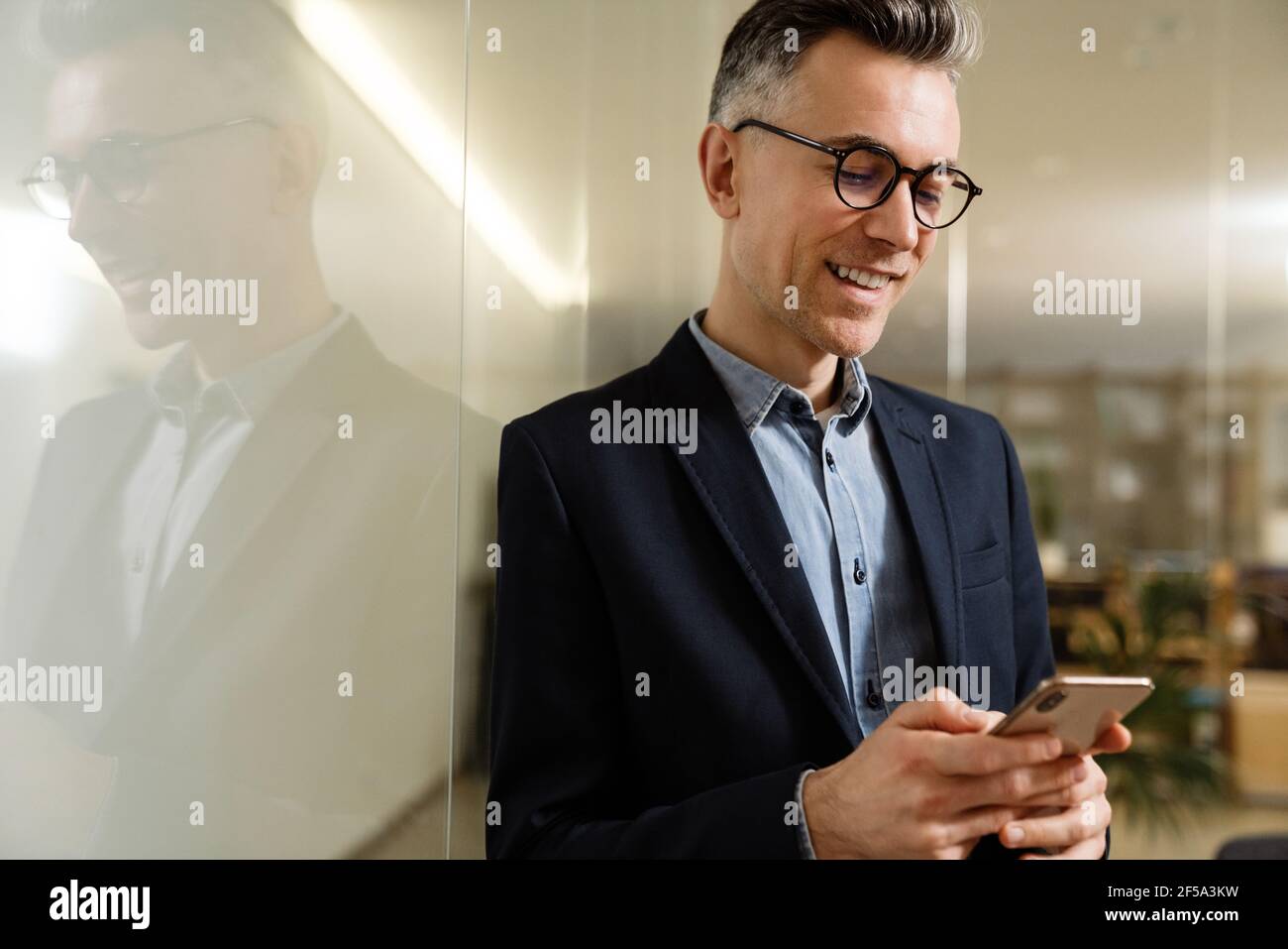Smiling grey man using mobile phone while leaning on wall in office ...