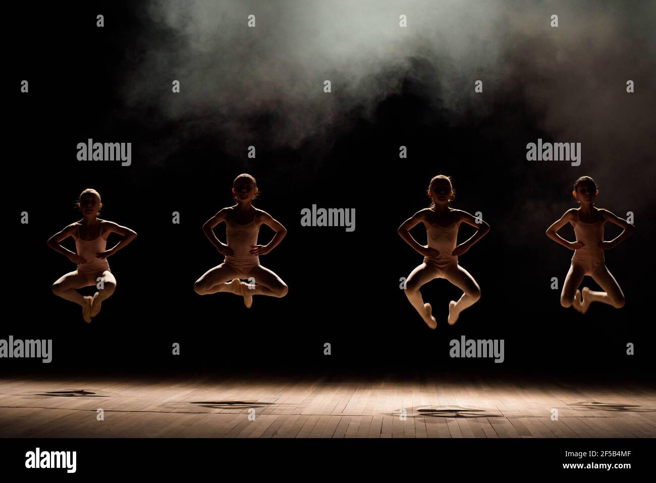 A group of small ballet dancers rehearses on stage with light and smoke ...