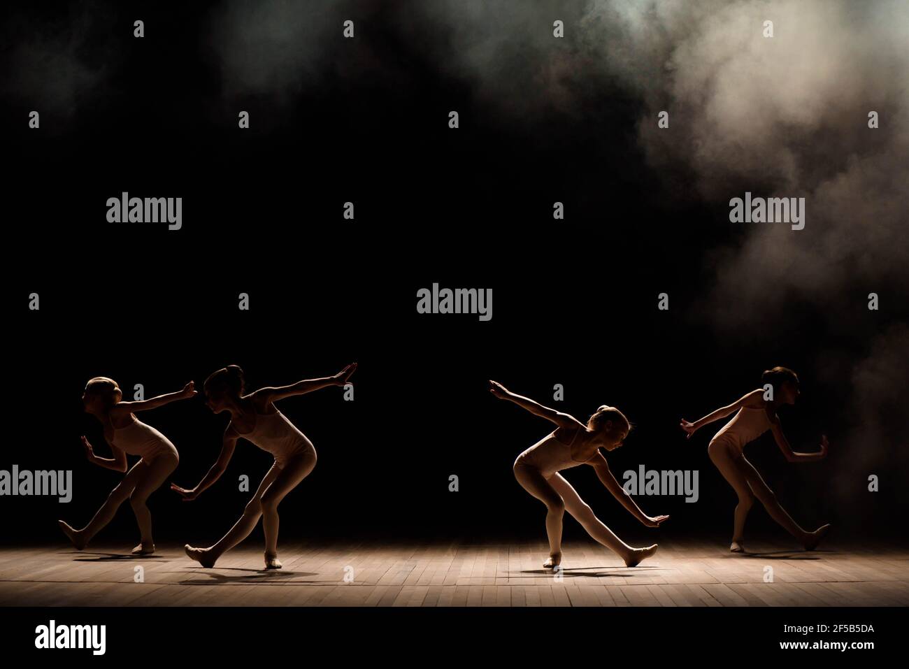 A group of small ballet dancers rehearses on stage with light and smoke ...