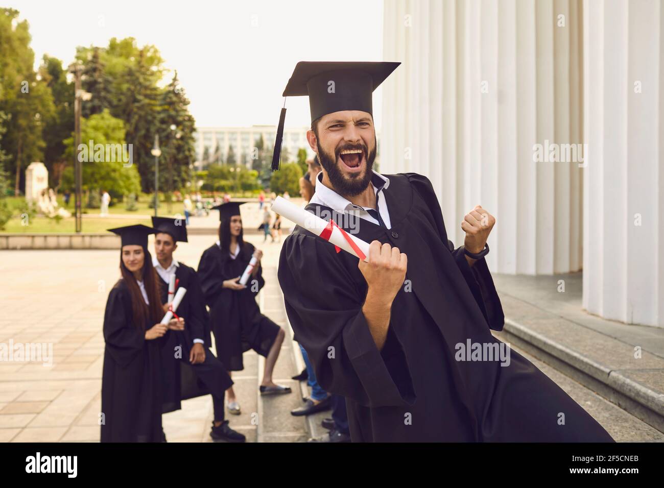 Emotional graduate wearing masters cap and gown raising up his hand ...