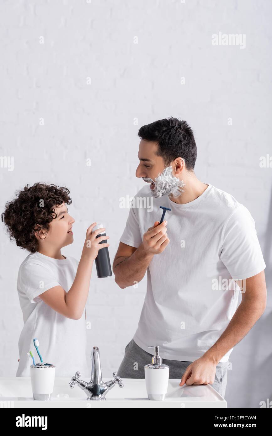 Smiling muslim boy holding shaving foam near father with razor and sink ...