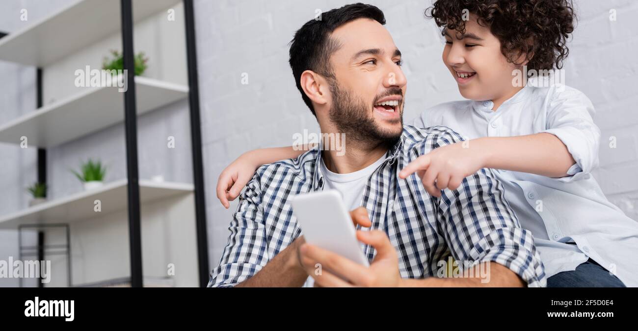 cheerful muslim boy pointing at smartphone in hands of smiling father ...