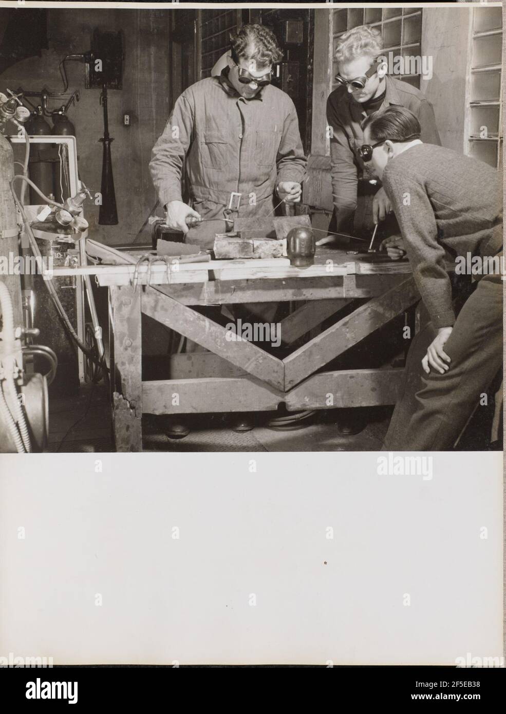 Three men wearing goggles weld a piece of metal on a table. Arnold ...