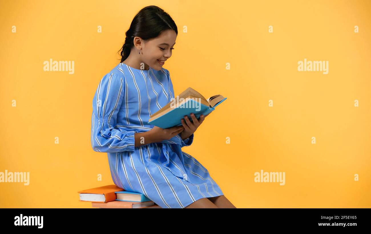 happy schoolgirl in blue dress reading book while sitting on yellow ...