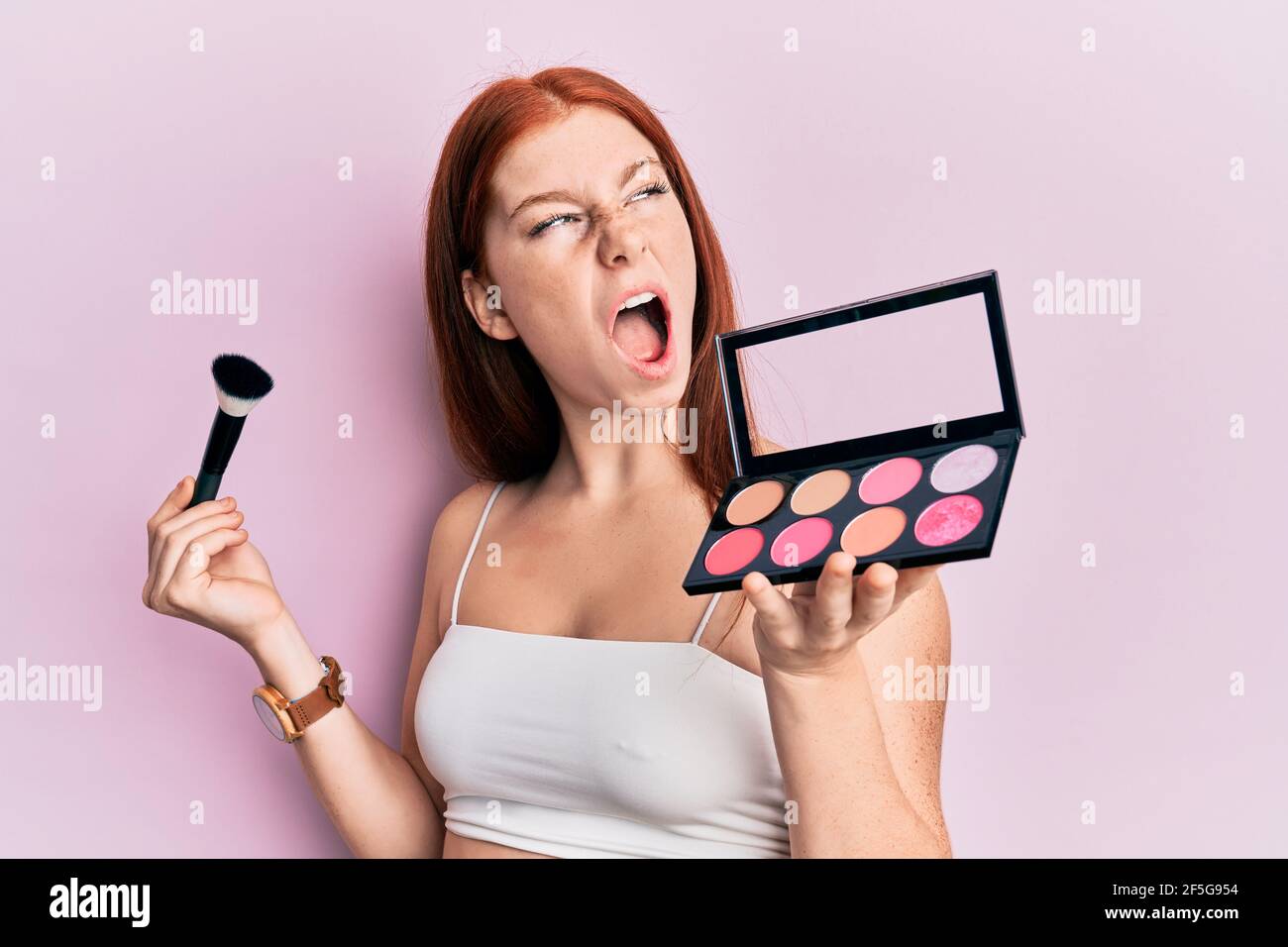 Young red head girl holding makeup brush and blush palette angry and ...