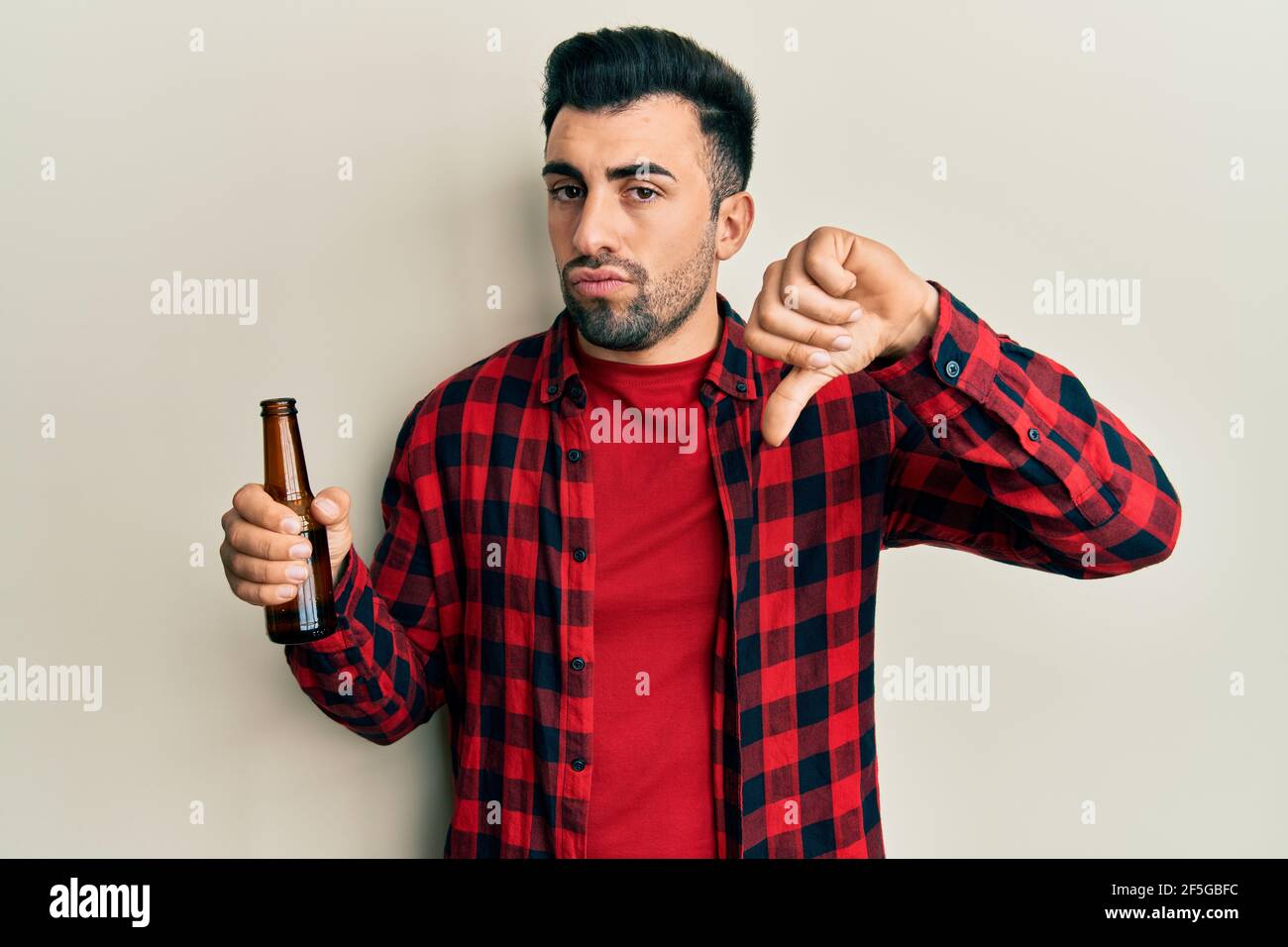 Young hispanic man drinking a bottle of beer with angry face, negative ...