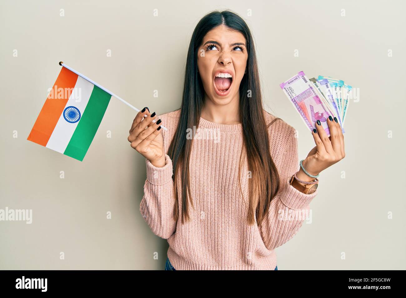 Young hispanic woman holding india flag and rupee banknotes angry and ...