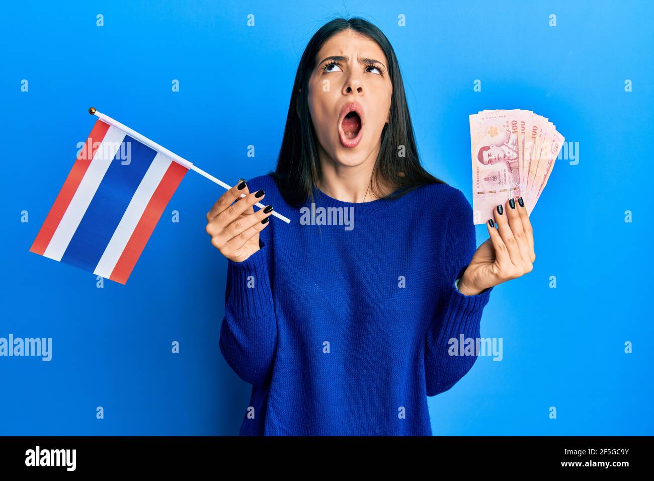 Young hispanic woman holding thailand flag and baht banknotes angry and ...