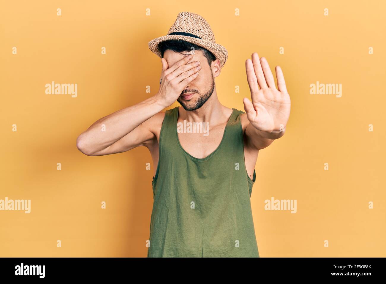 Young hispanic man wearing summer hat covering eyes with hands and ...