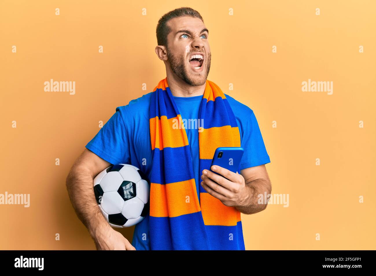 Young caucasian man holding football ball looking at smartphone angry ...