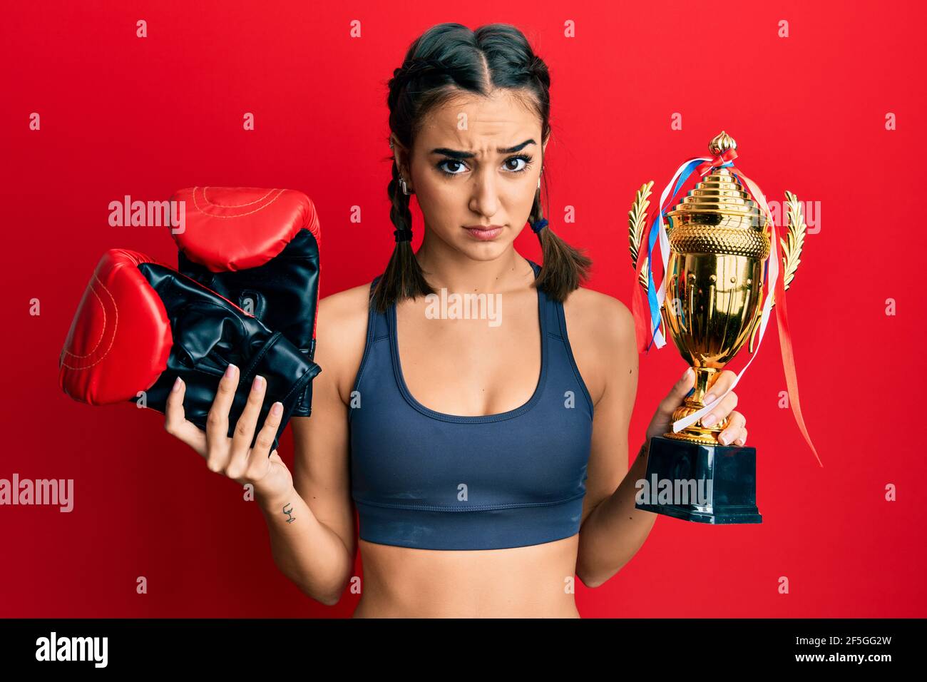 Young brunette girl holding boxer gloves and trophy skeptic and nervous ...
