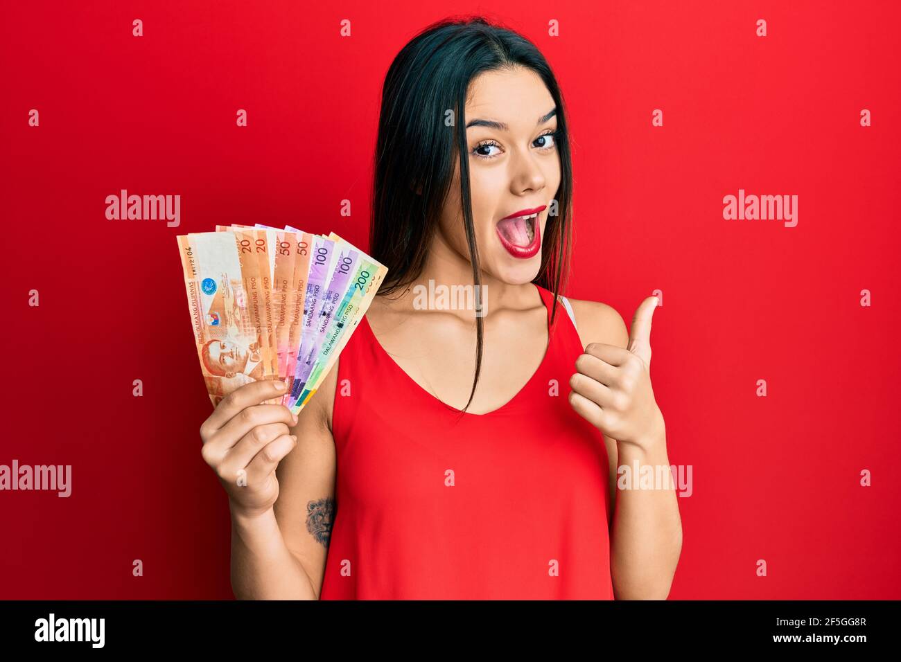 Young hispanic girl holding philippine peso banknotes pointing thumb up ...