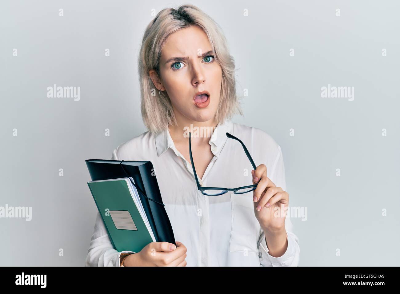 Young blonde girl reading a book and holding glasses in shock face ...